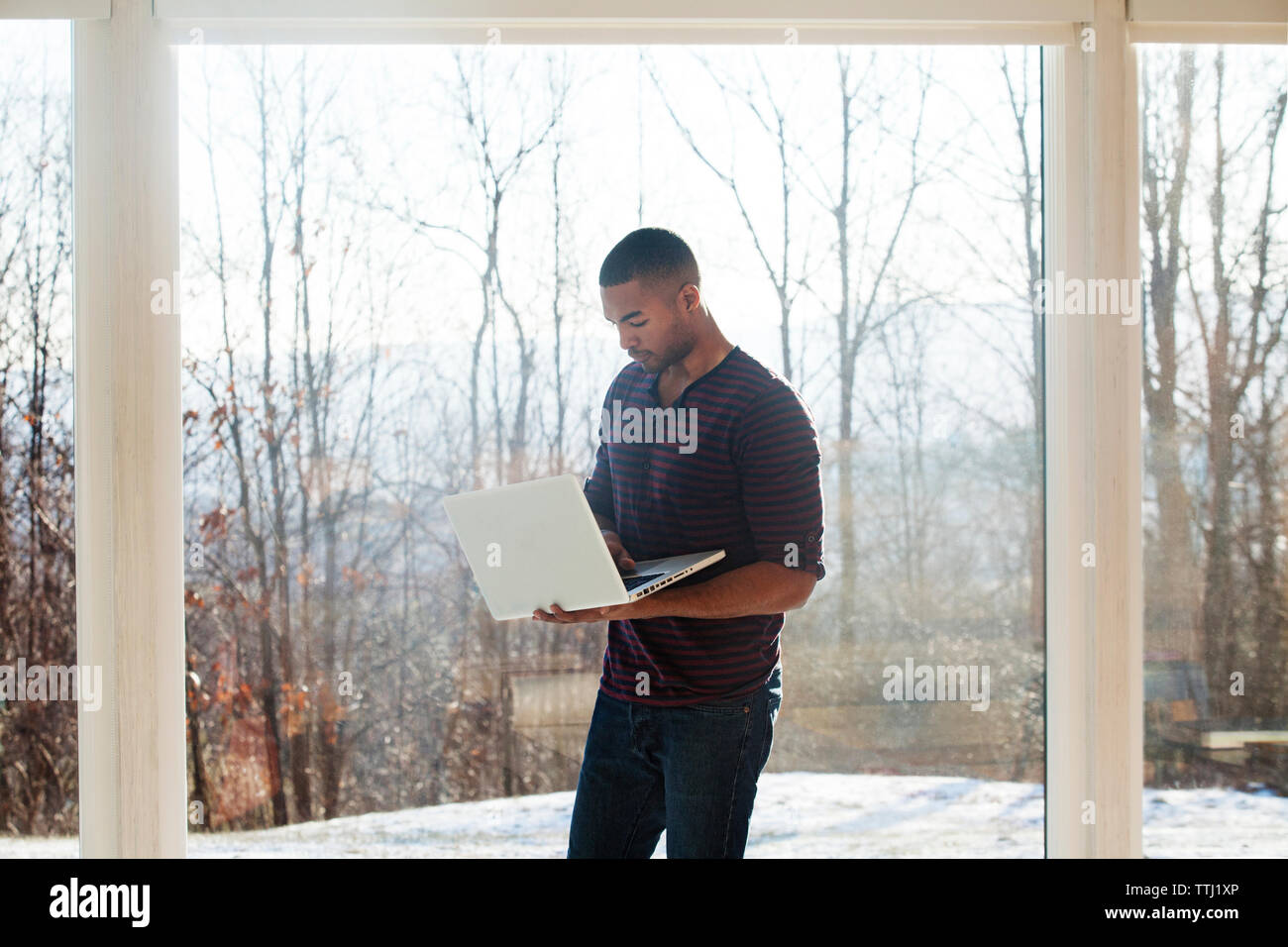 Man using laptop computer while standing against glass window at home ...