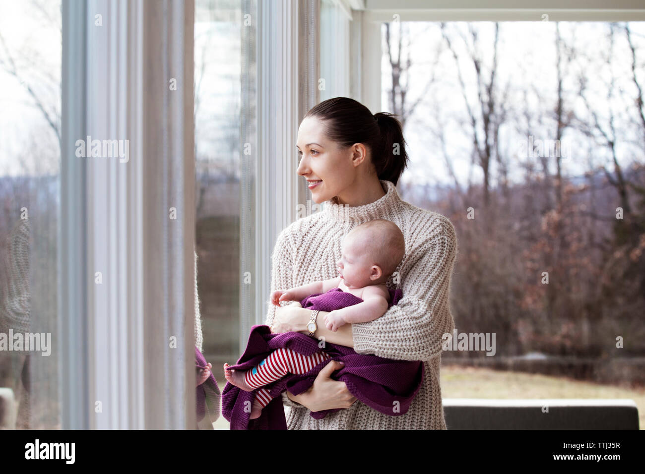 Thoughtful woman carrying baby girl while looking through window at ...