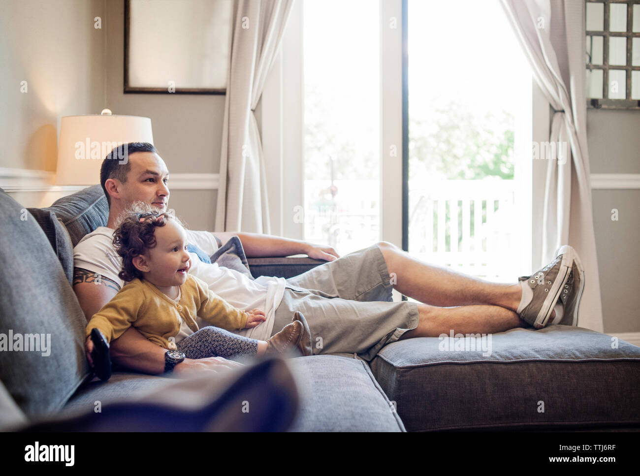 Father and daughter watching television while sitting on sofa at home ...