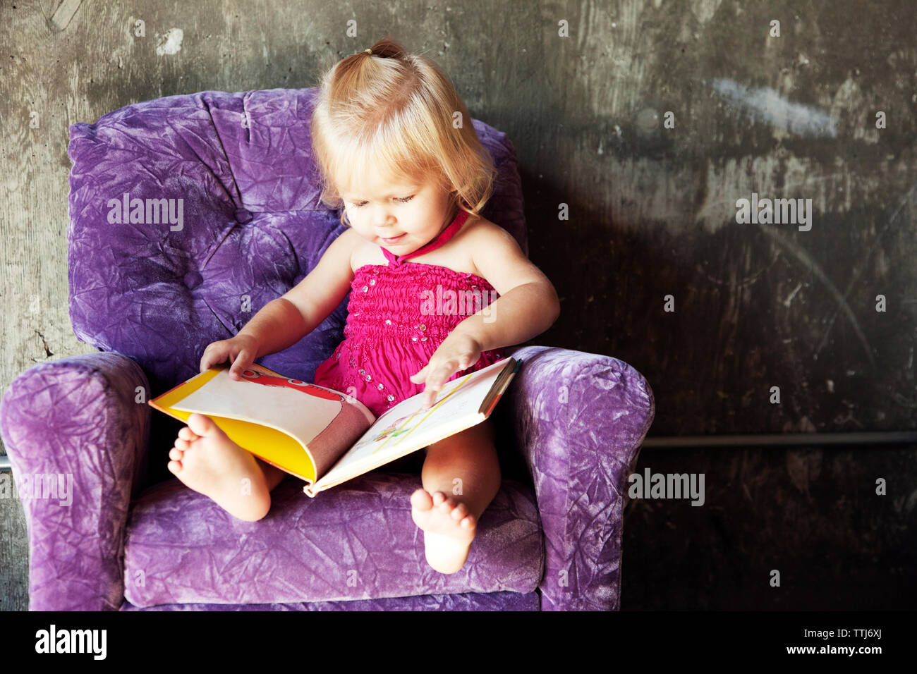 Baby girl looking picture book while sitting on arm chair at home Stock ...