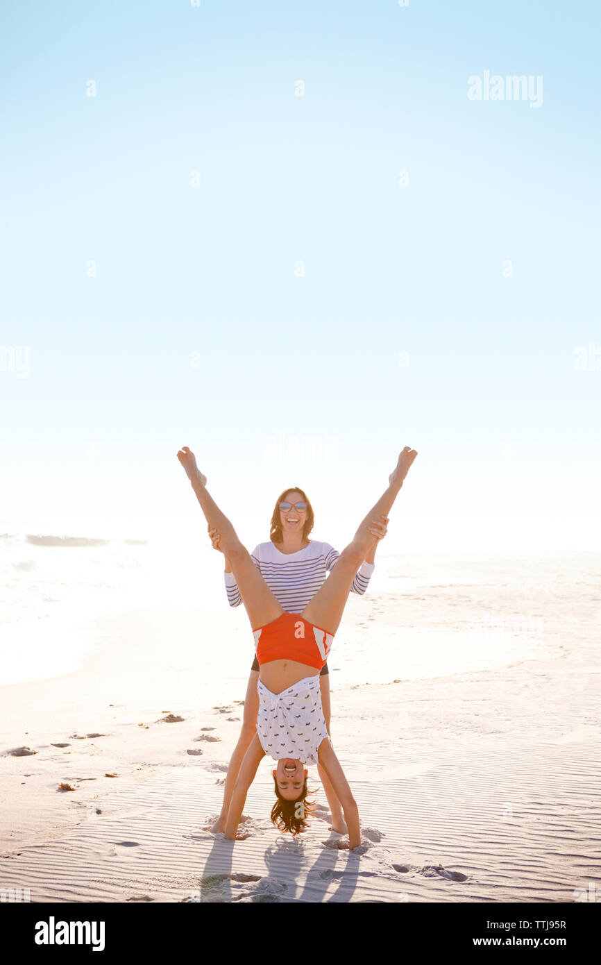 Portrait of woman holding legs of friend doing hand stand on sand at ...