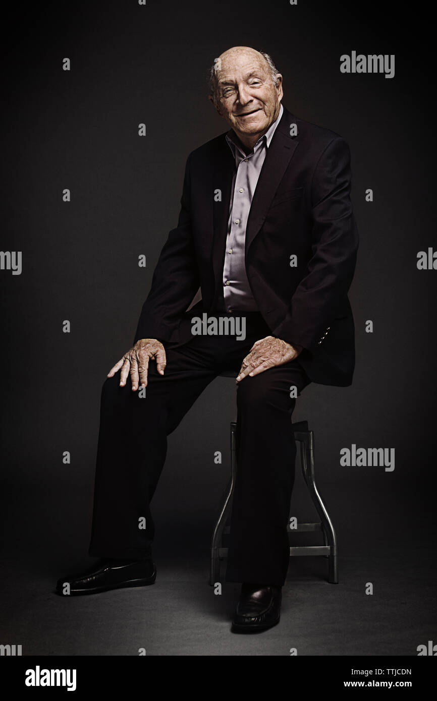 Portrait of senior man sitting on stool against black background Stock ...