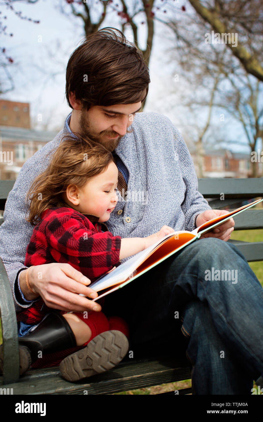 Father and daughter reading book while sitting on bench at park Stock ...
