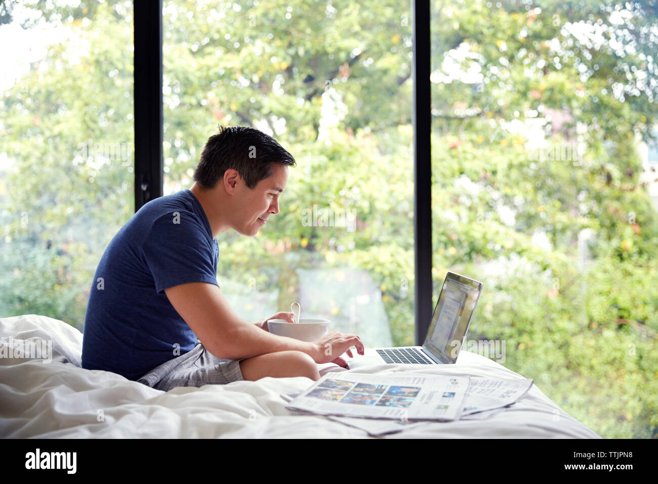 Side view of man using laptop while having breakfast on bed by window ...