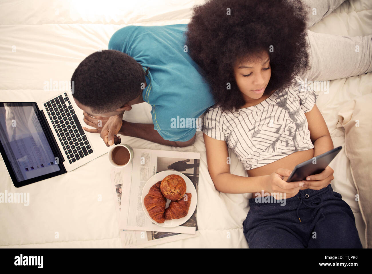 Couple having croissants and using technology while resting on bed at ...