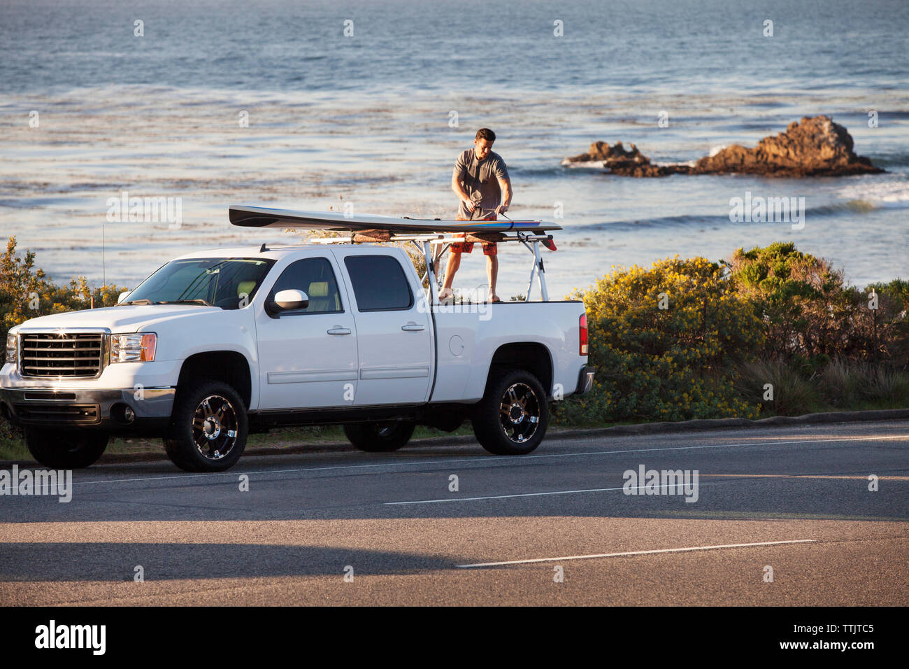Man tying surfboard on pick-up truck parked at roadside against sea ...