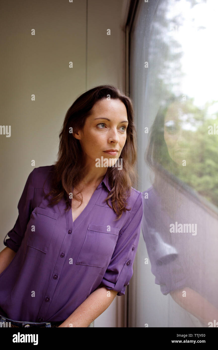 Thoughtful woman looking through window while leaning against wall at ...