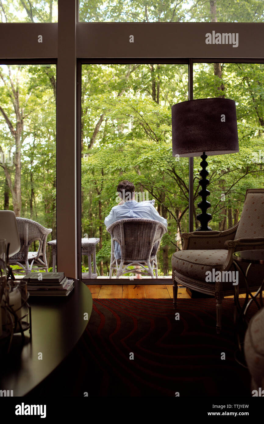 Rear view of man sitting on chair at porch seen through window from ...