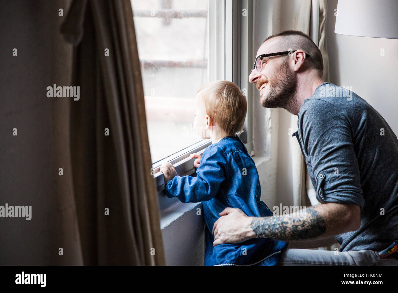 Happy father with daughter looking through window while sitting at home ...