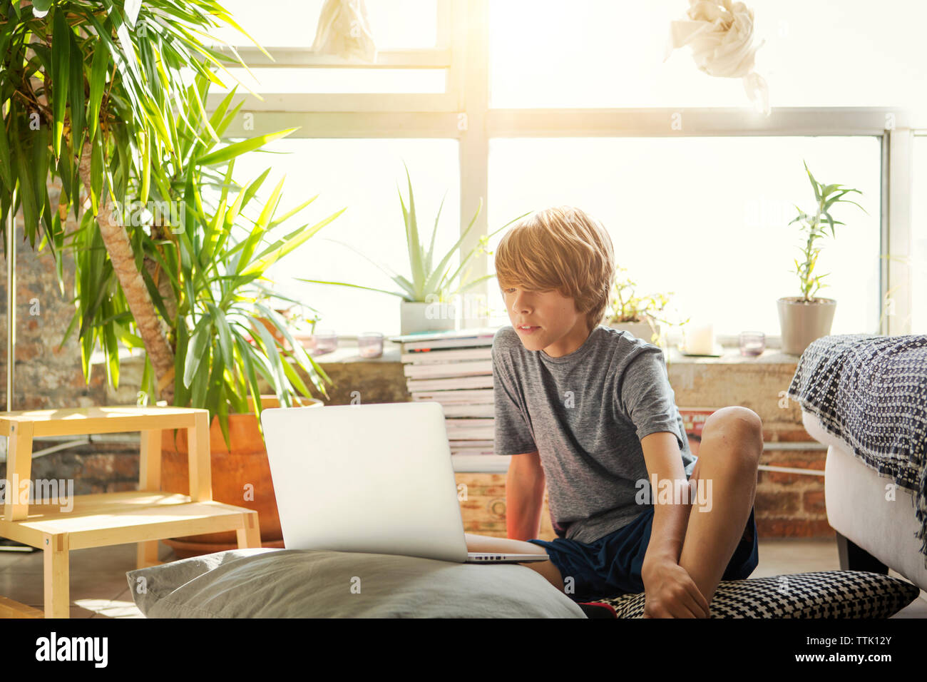 Boy looking at laptop computer while sitting on floor against window ...