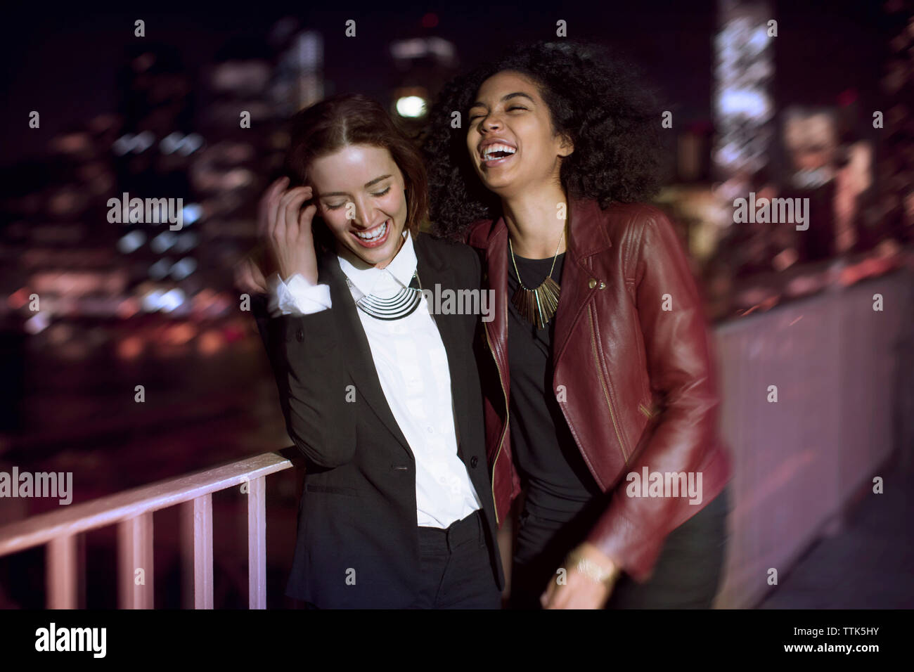 Female friends laughing while standing by railing on street at night ...