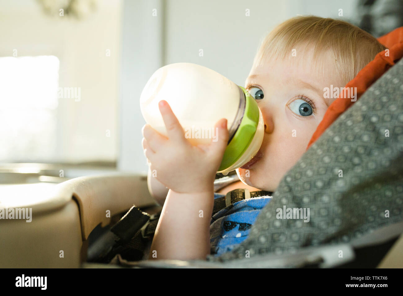 Portrait of baby boy drinking milk while sitting on high chair at home ...