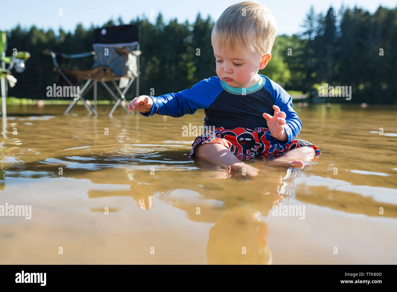Cute baby boy playing with water while sitting in lake against trees ...