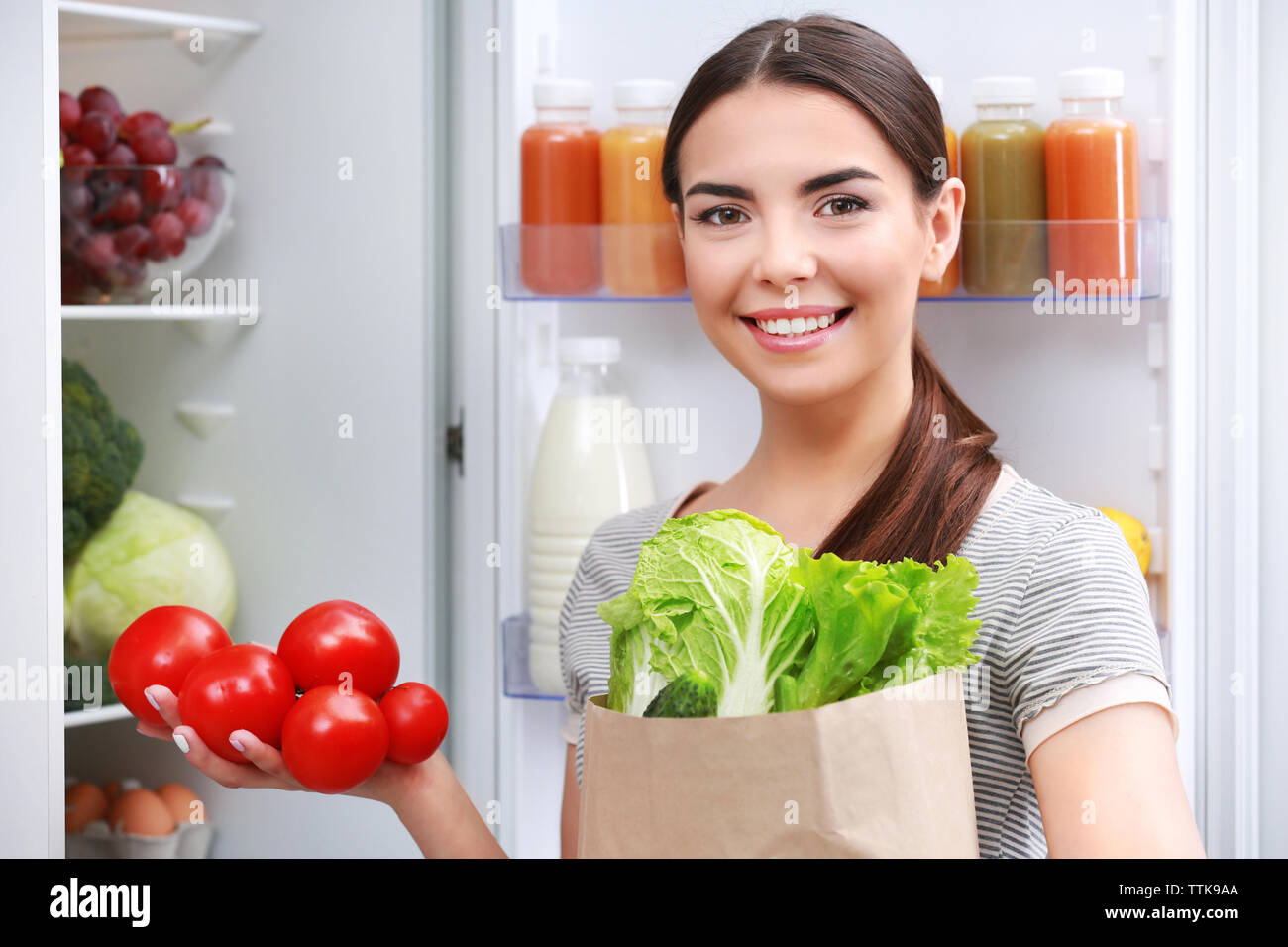 Young woman with purchase box full of vegetables standing beside fridge ...