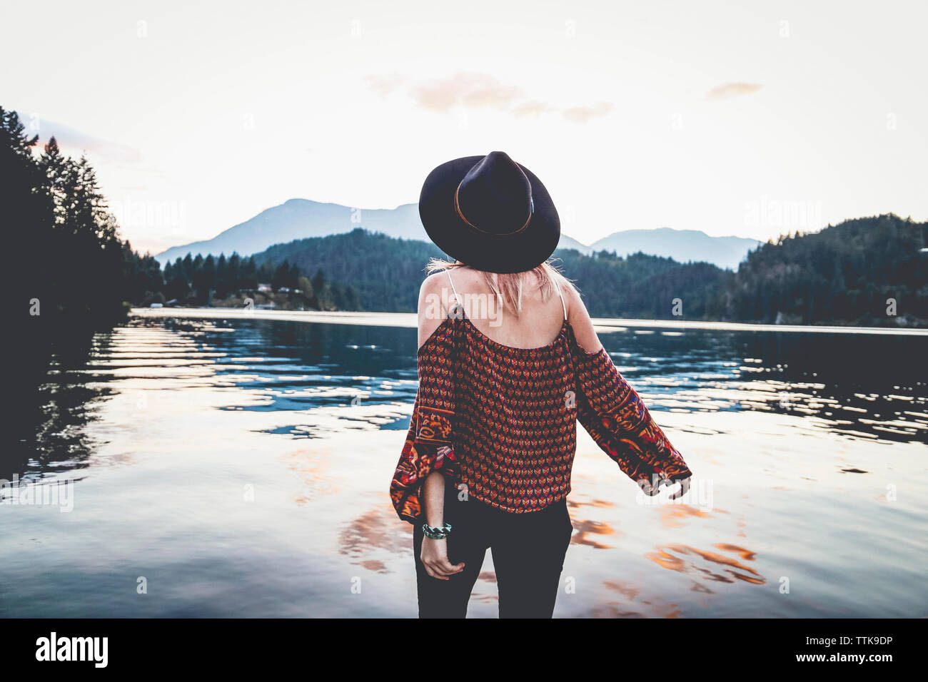 Rear view of woman if fedora hat looking at view while standing by lake ...