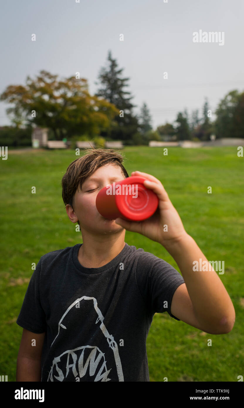 Boy drinking water while standing on grassy field against sky in park ...