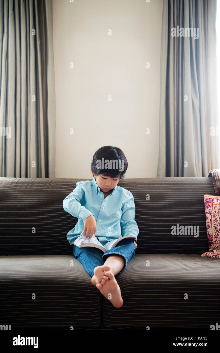 Boy reading book while sitting on sofa against wall at home Stock Photo ...