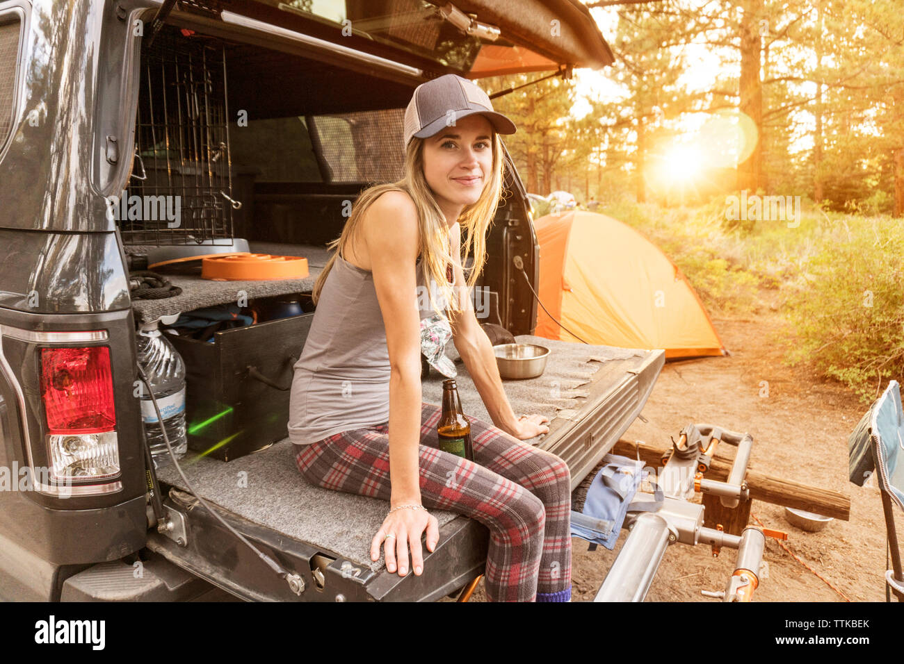 Portrait of smiling woman with beer bottle sitting on car trunk against ...