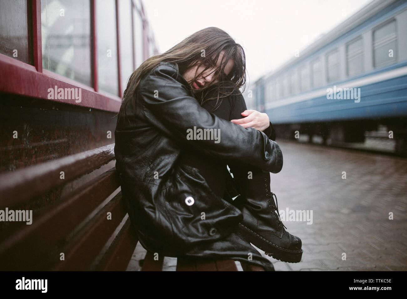 Portrait of sad woman sitting on bench at railroad station during rainy ...
