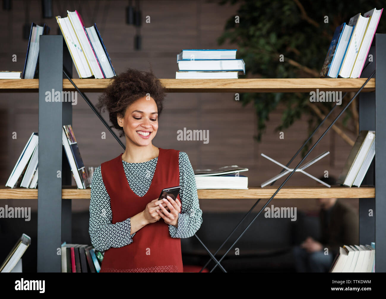 Happy businesswoman using smart phone while standing against book shelf ...