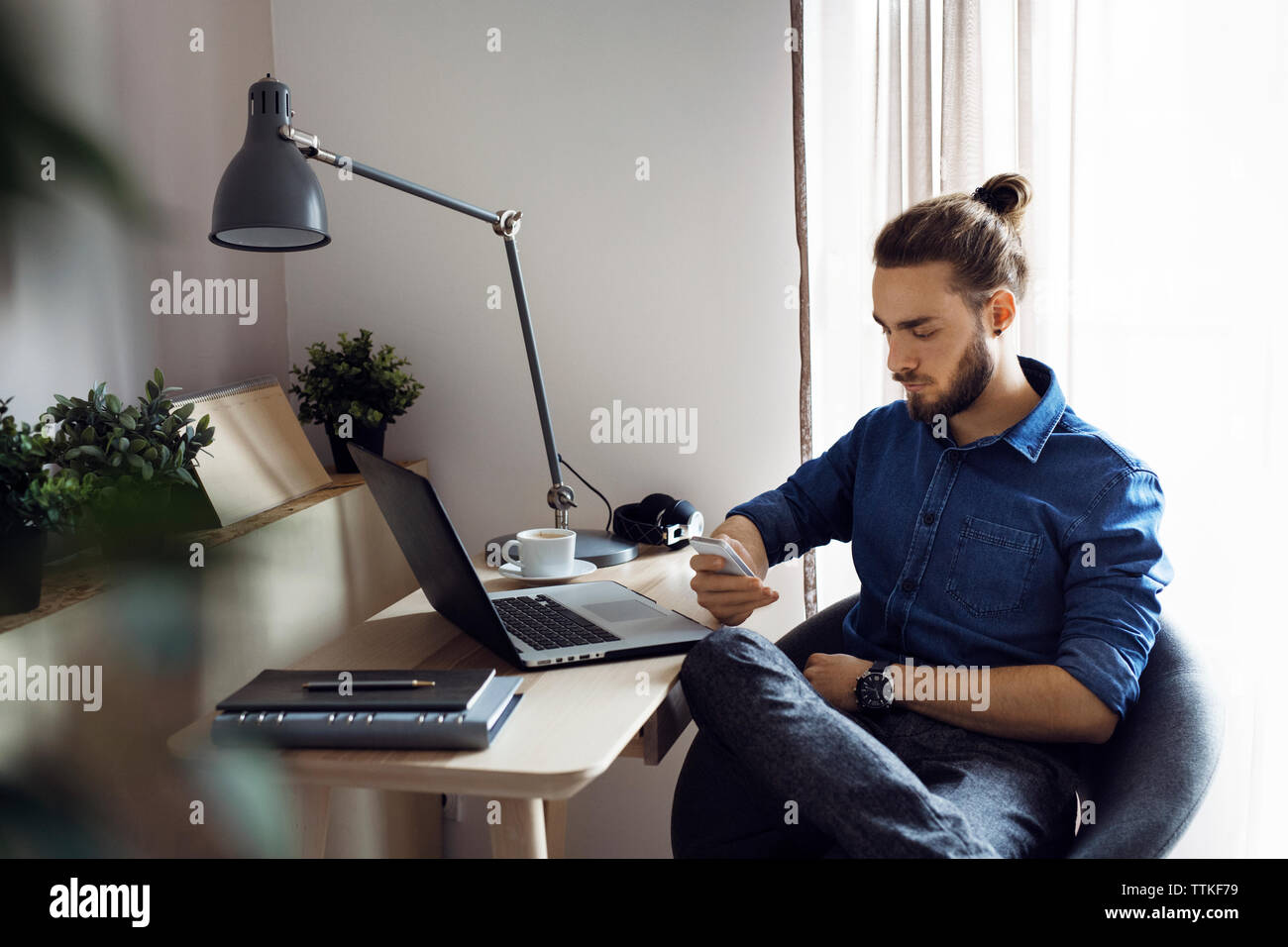 Man using mobile phone while sitting on chair by window at home Stock ...