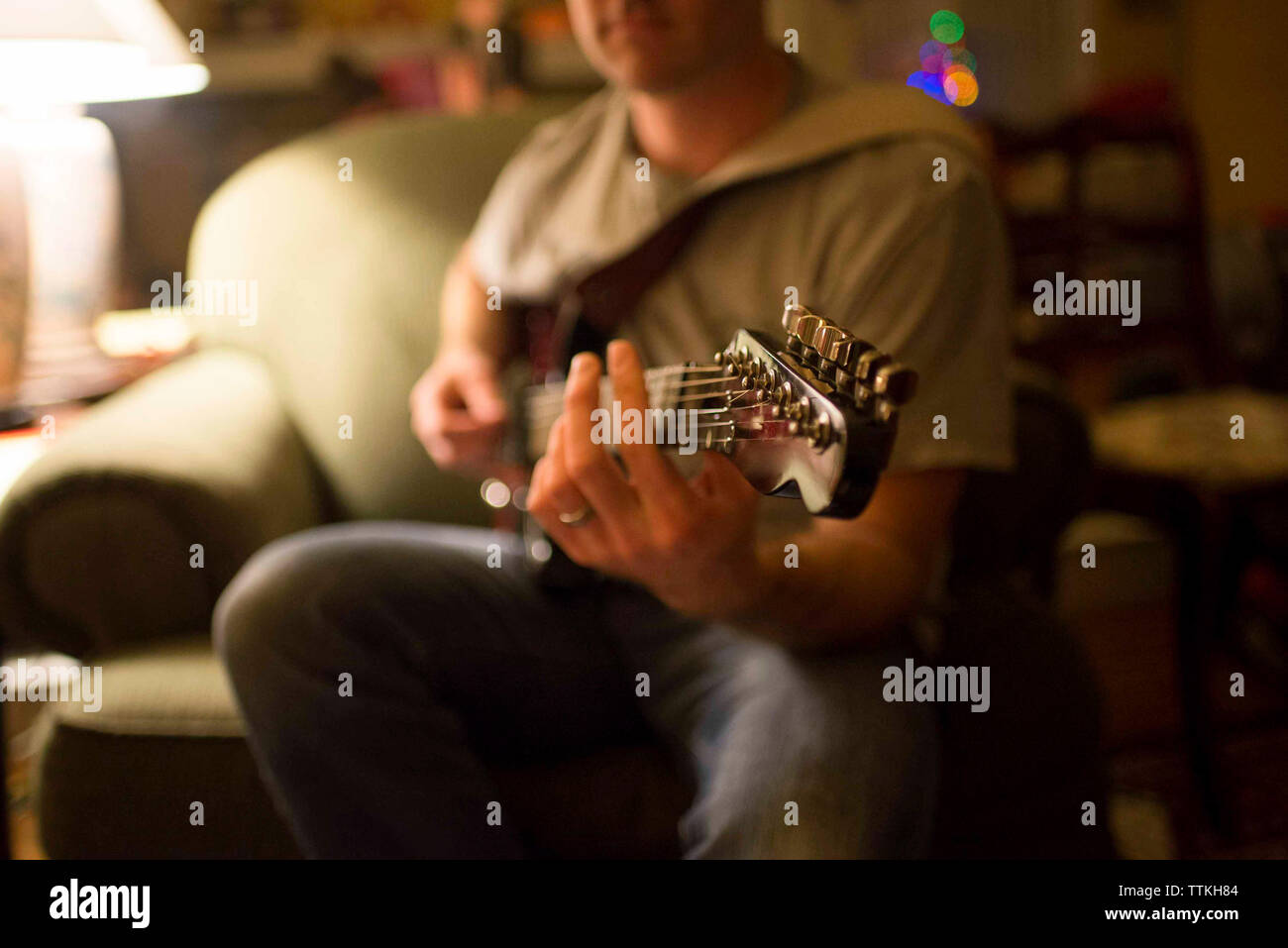 Midsection of man playing guitar while sitting on armchair at home ...