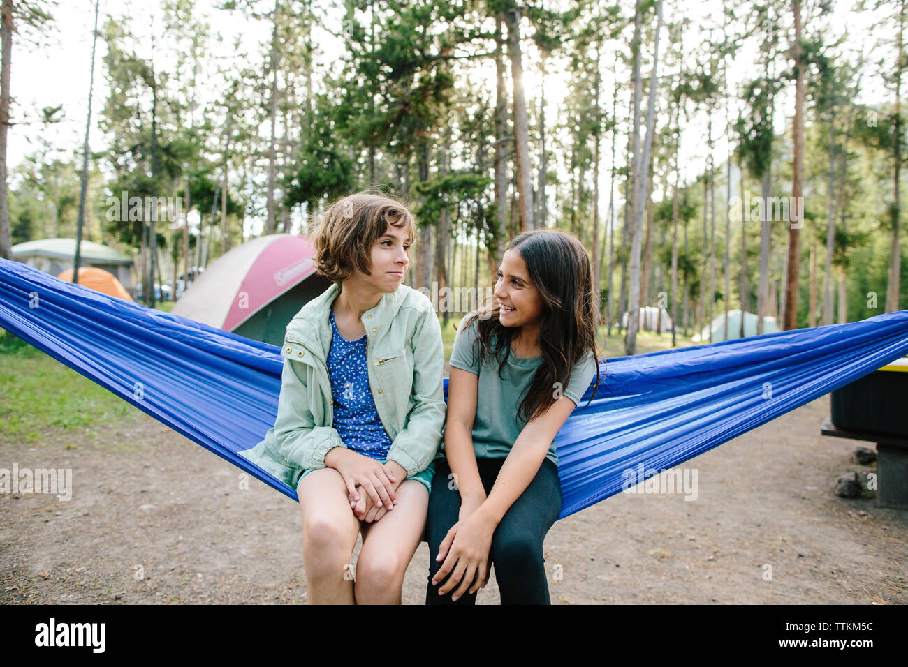 Sisters Talking While Sitting On Hammock Against Trees In Forest Stock