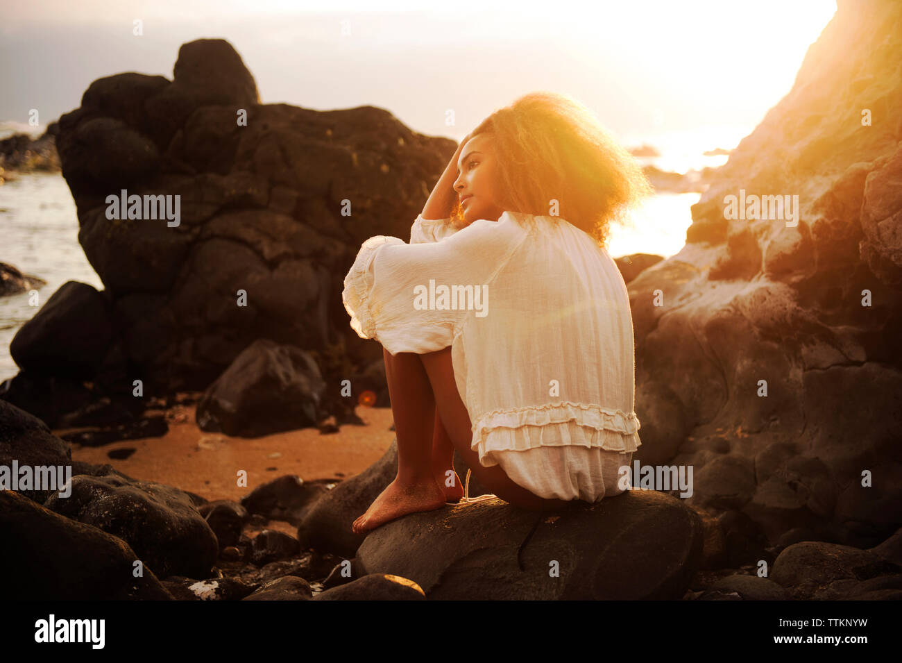 Thoughtful teenage girl sitting on rock at seashore during sunny day ...