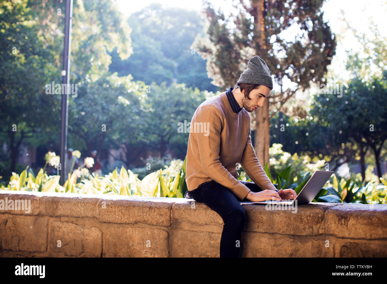 Side view of man using laptop computer while sitting on retaining wall ...