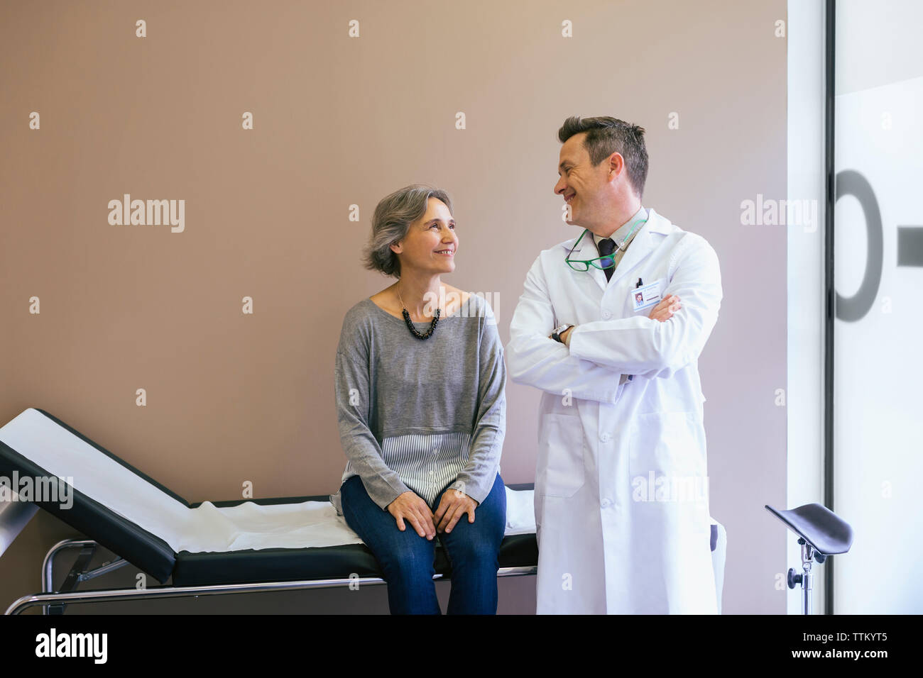 Smiling patient looking at doctor while sitting on bed against wall at ...