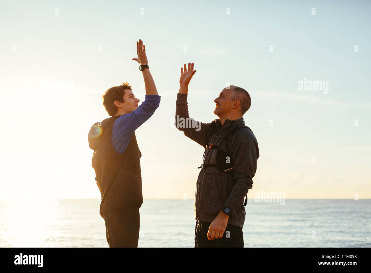 Happy father and son having high-five against sea and sky during sunset ...