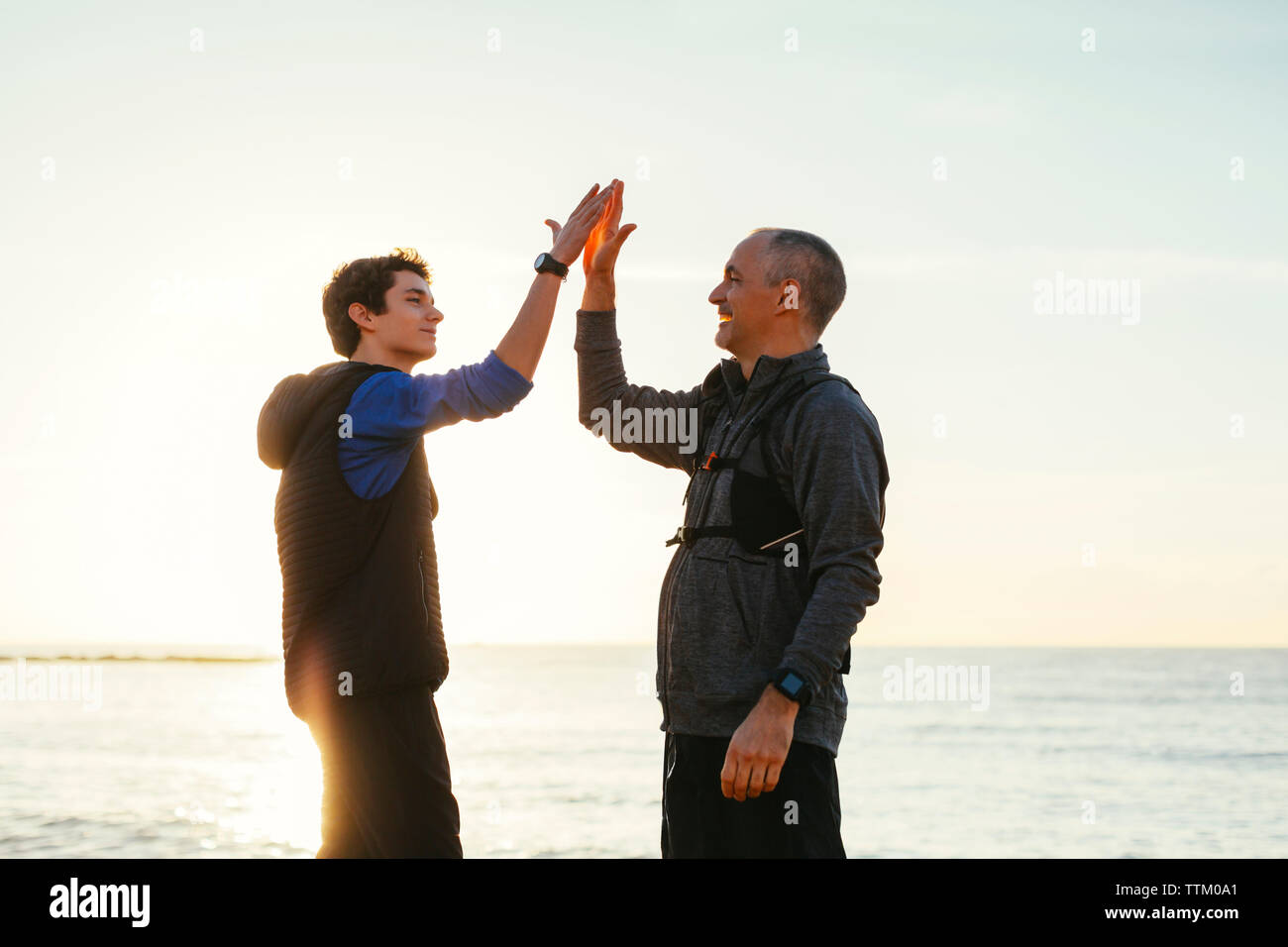 Happy father and son doing high-five against sea and sky during sunset ...