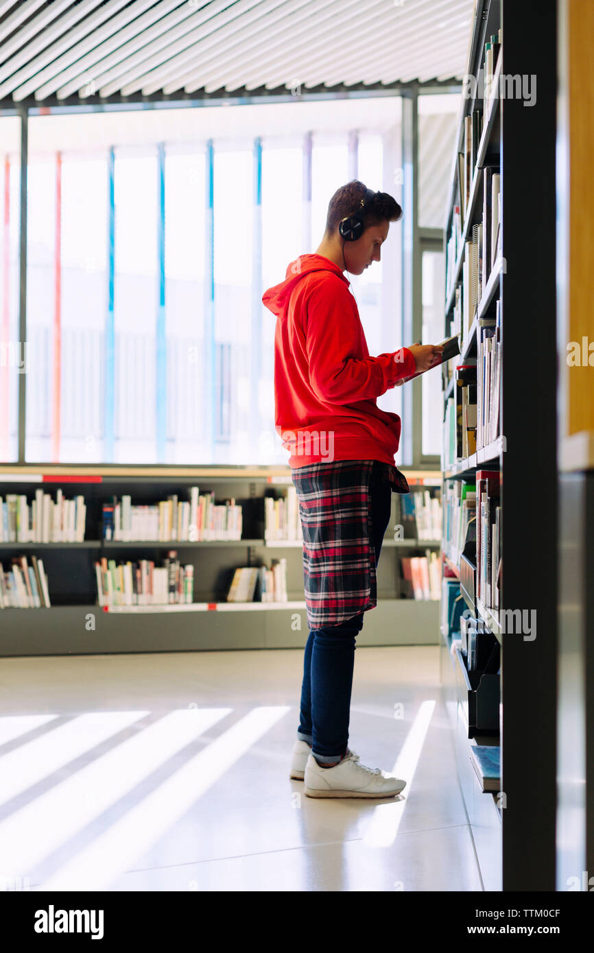 Side view of man with headphones reading book while standing in library ...