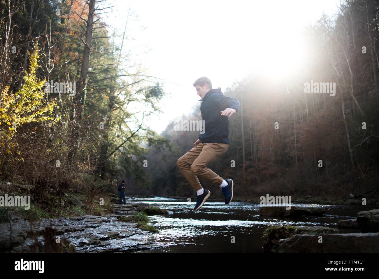 Side view of teenage boy jumping over river against trees at forest ...