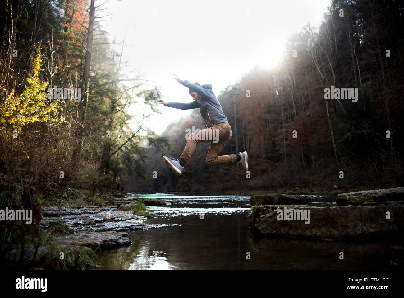 Side view of teenage boy jumping over river against trees in forest ...