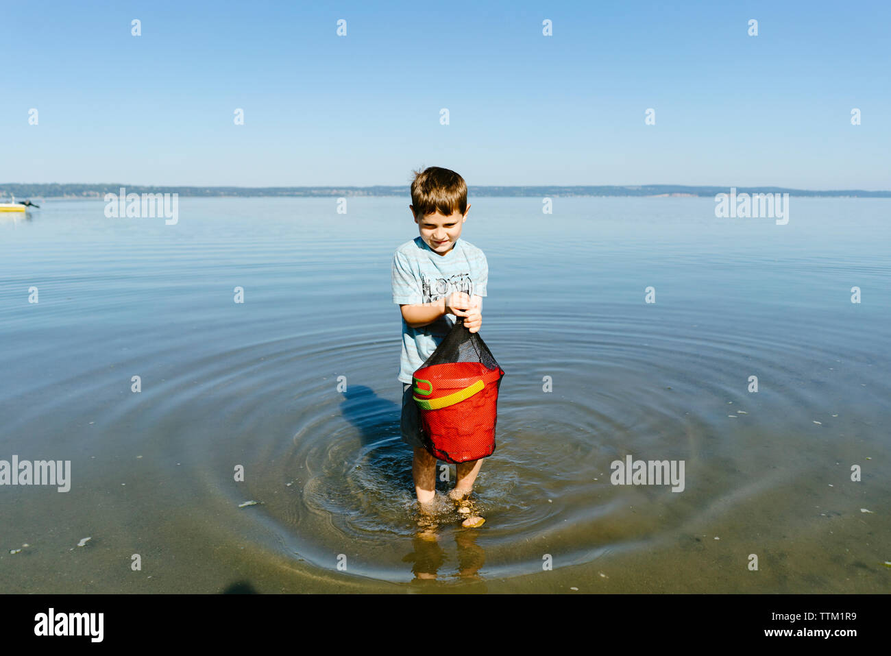 Boy carrying bucket while standing in sea against clear blue sky during ...