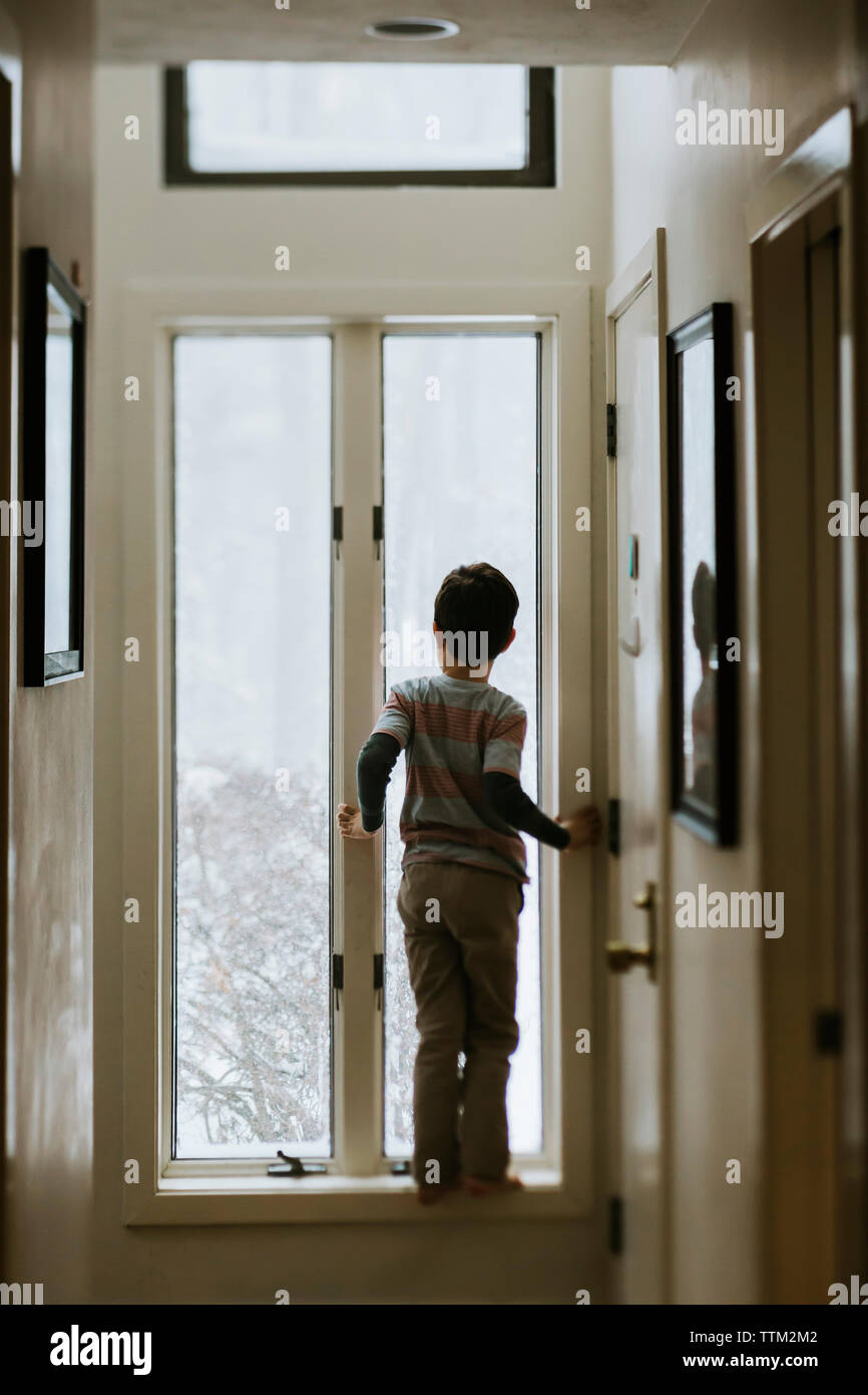 Rear view of boy looking through window while standing at home Stock ...