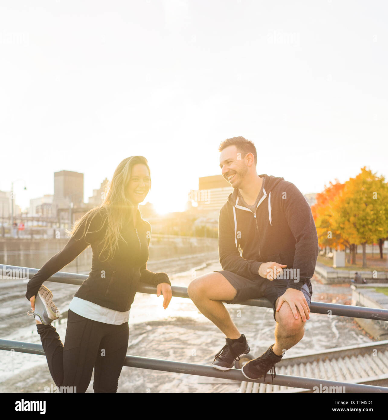 Happy man looking at woman stretching while sitting on railing against ...