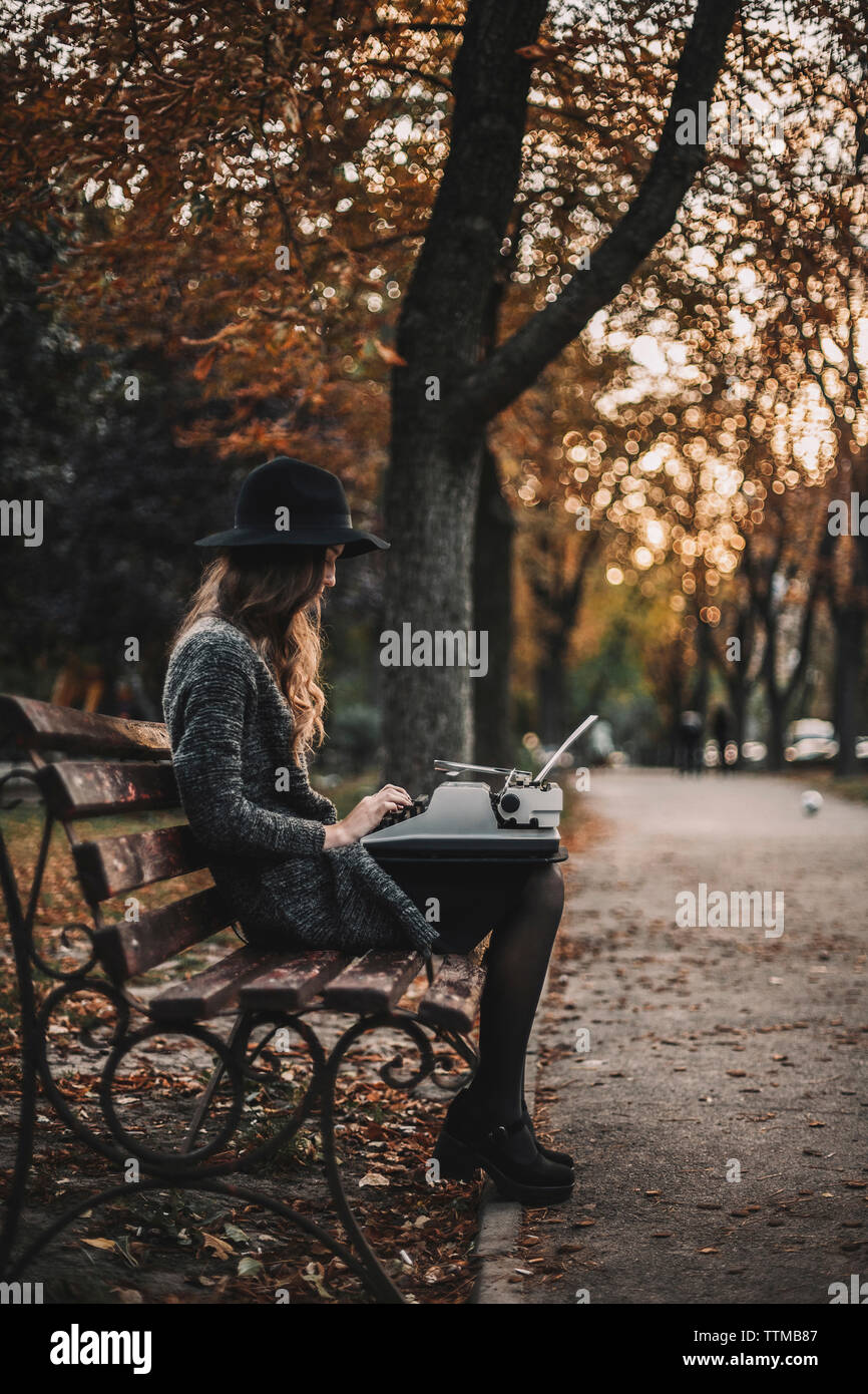Side view of writer using typewriter while sitting on bench at park ...