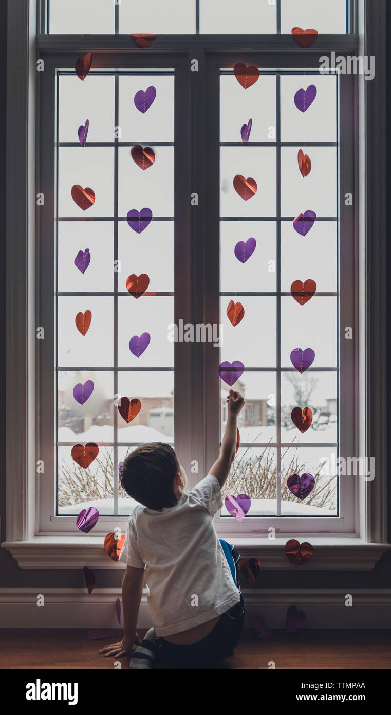 Rear view of boy playing with heart shape decorations by window at home ...