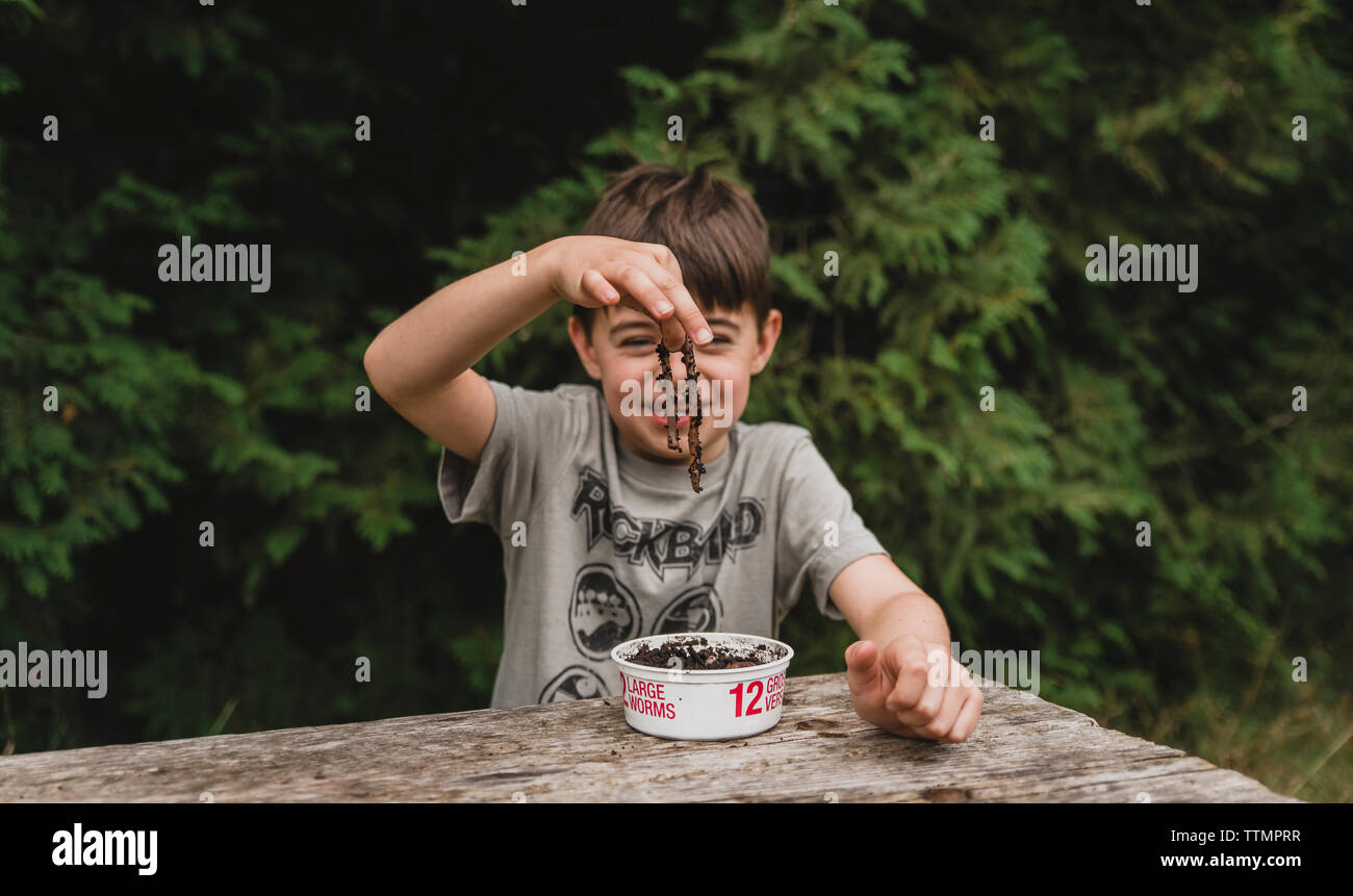 Boy holding earthworm at wooden table while sitting against trees in ...