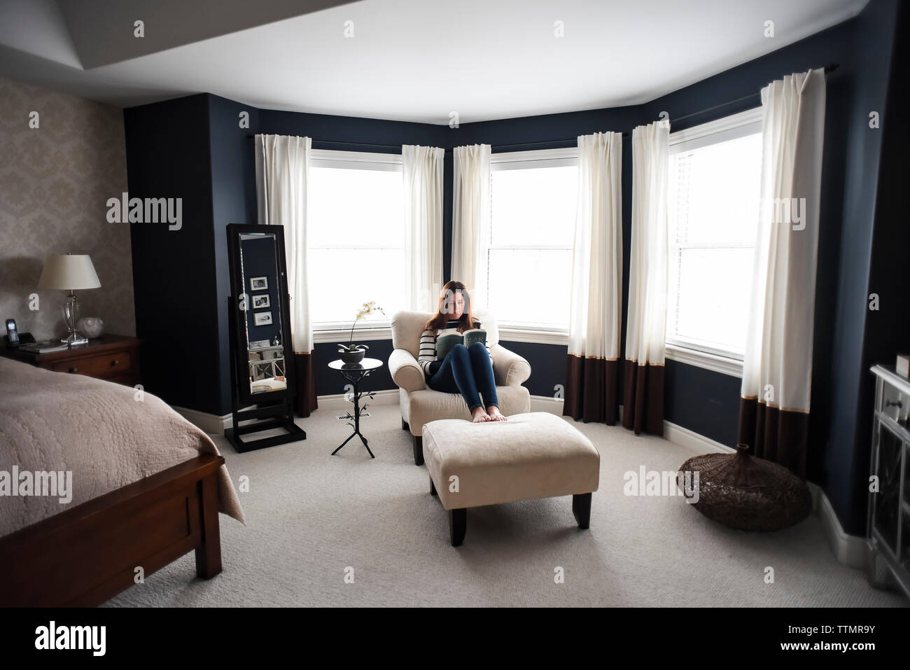 Woman sitting in chair reading book in front of windows in a bedroom ...