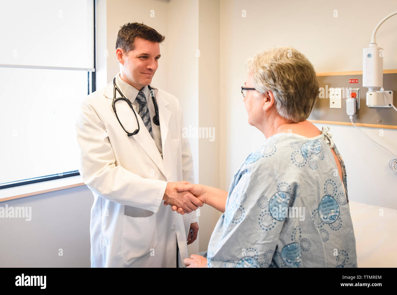 Doctor shaking hands with older patient in gown in clinical setting ...