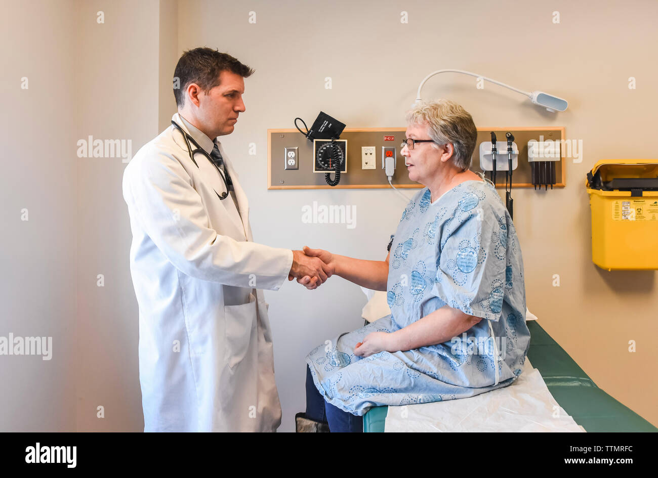 Doctor shaking hands with older patient in gown in clinical setting ...