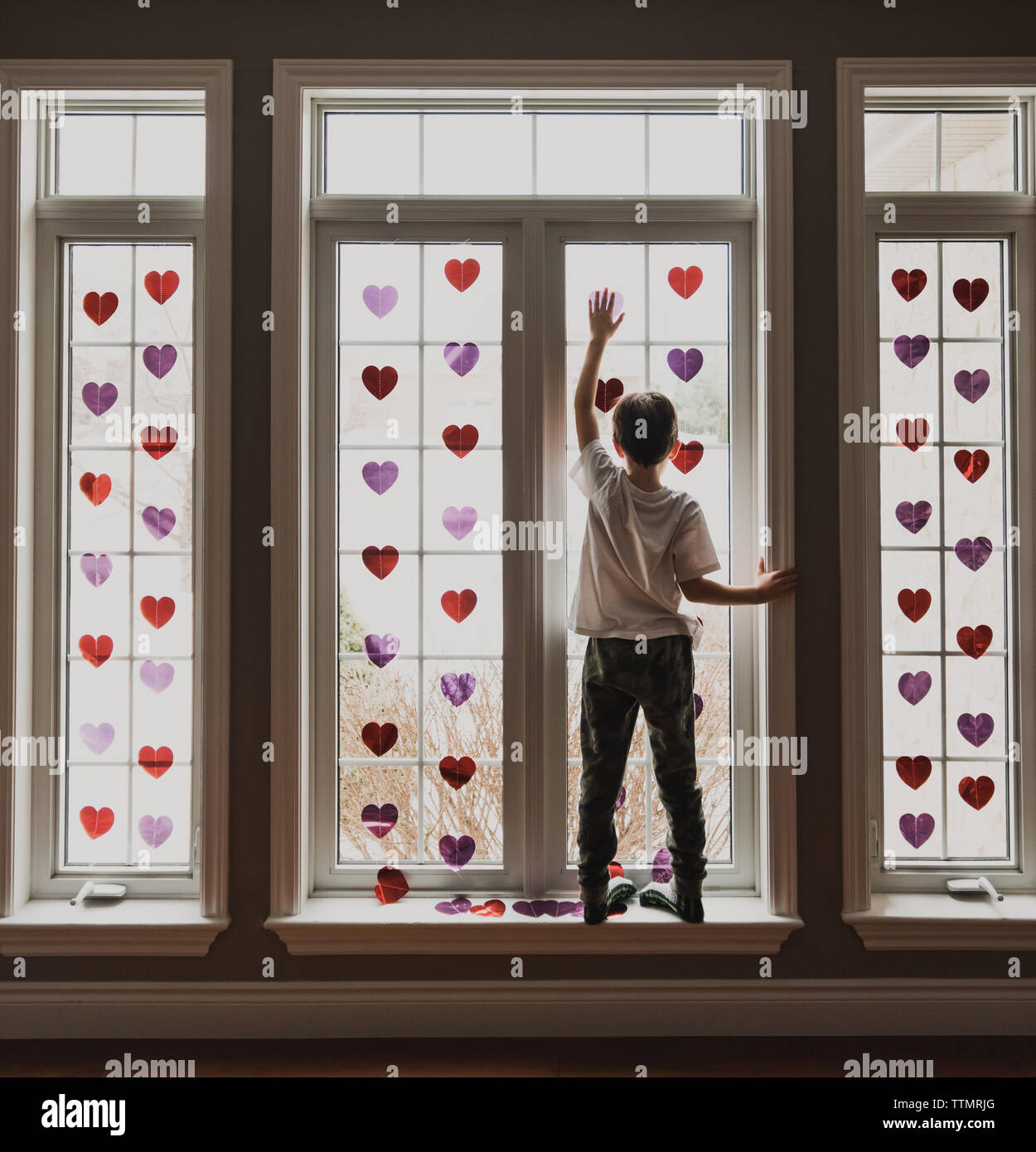 Young boy standing in window that is covered in small shiny hearts ...