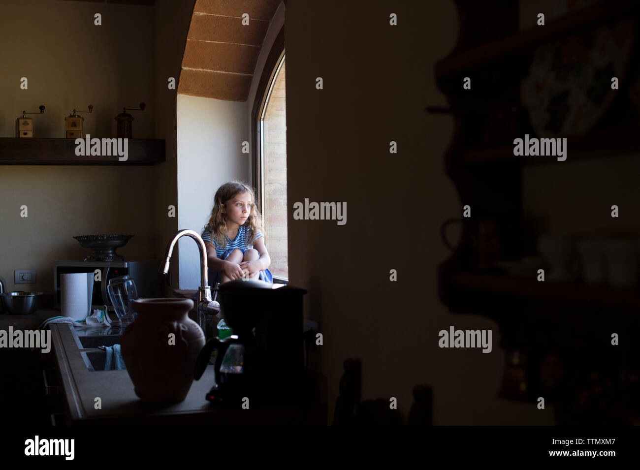 Girl looking through window while sitting in kitchen at home Stock ...
