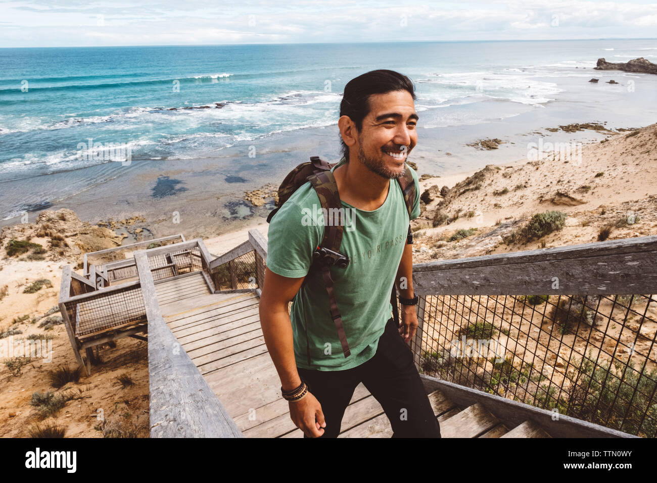 Smiling hiker with backpack climbing wooden steps at beach against sea ...