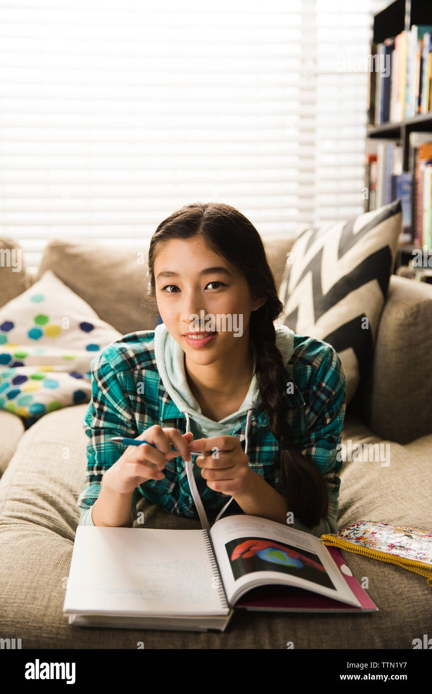 Portrait of teenage girl doing homework while lying on couch at home ...