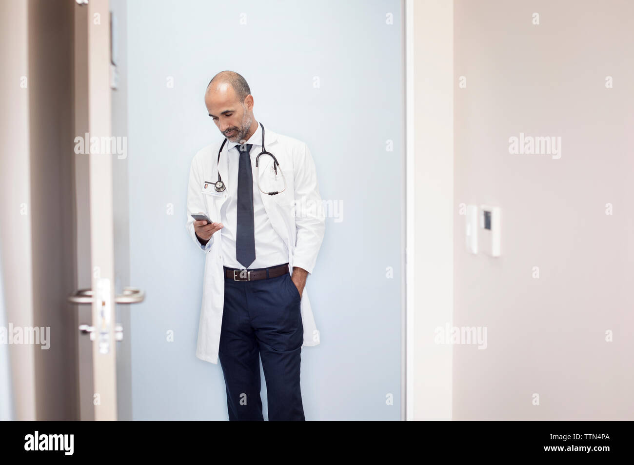 Doctor using smart phone while standing against wall at hospital seen ...