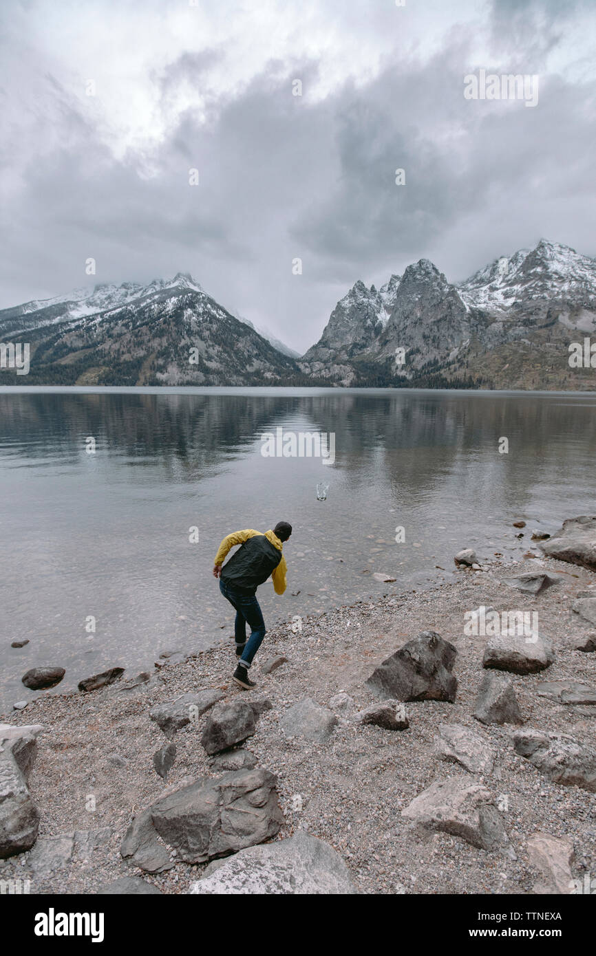 Full length of playful man throwing stone in lake against mountains and ...